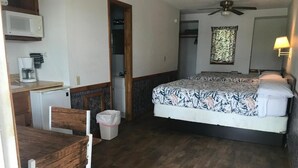 Quarto compacto com uma cama, pequeno espaço de cozinha, uma cadeira de madeira e ventilador de teto.
