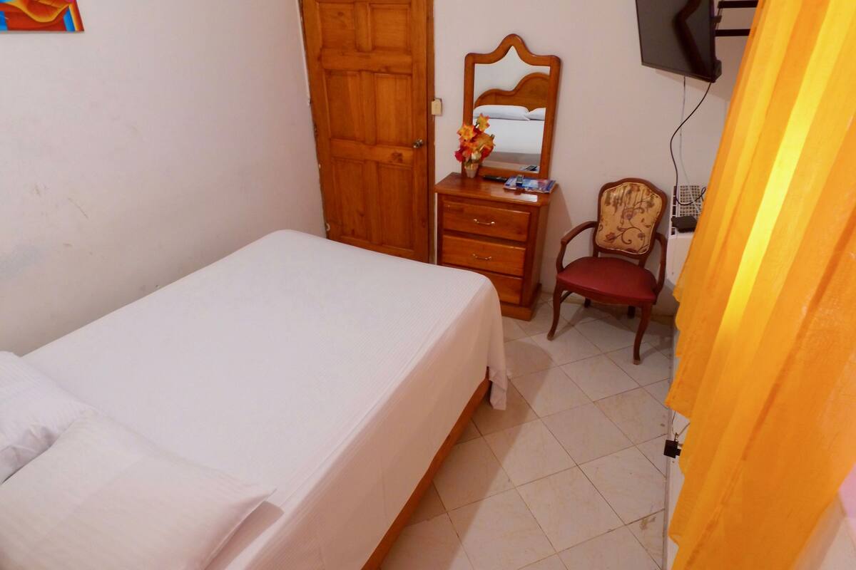 Quarto com cama, cômoda de madeira, cadeira e televisão fixada na parede.