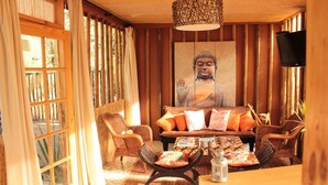 Um quarto com teto de madeira, pintura de Buda na parede, sofá com almofadas, cadeiras de vime, mesa com abajur e TV na parede.
