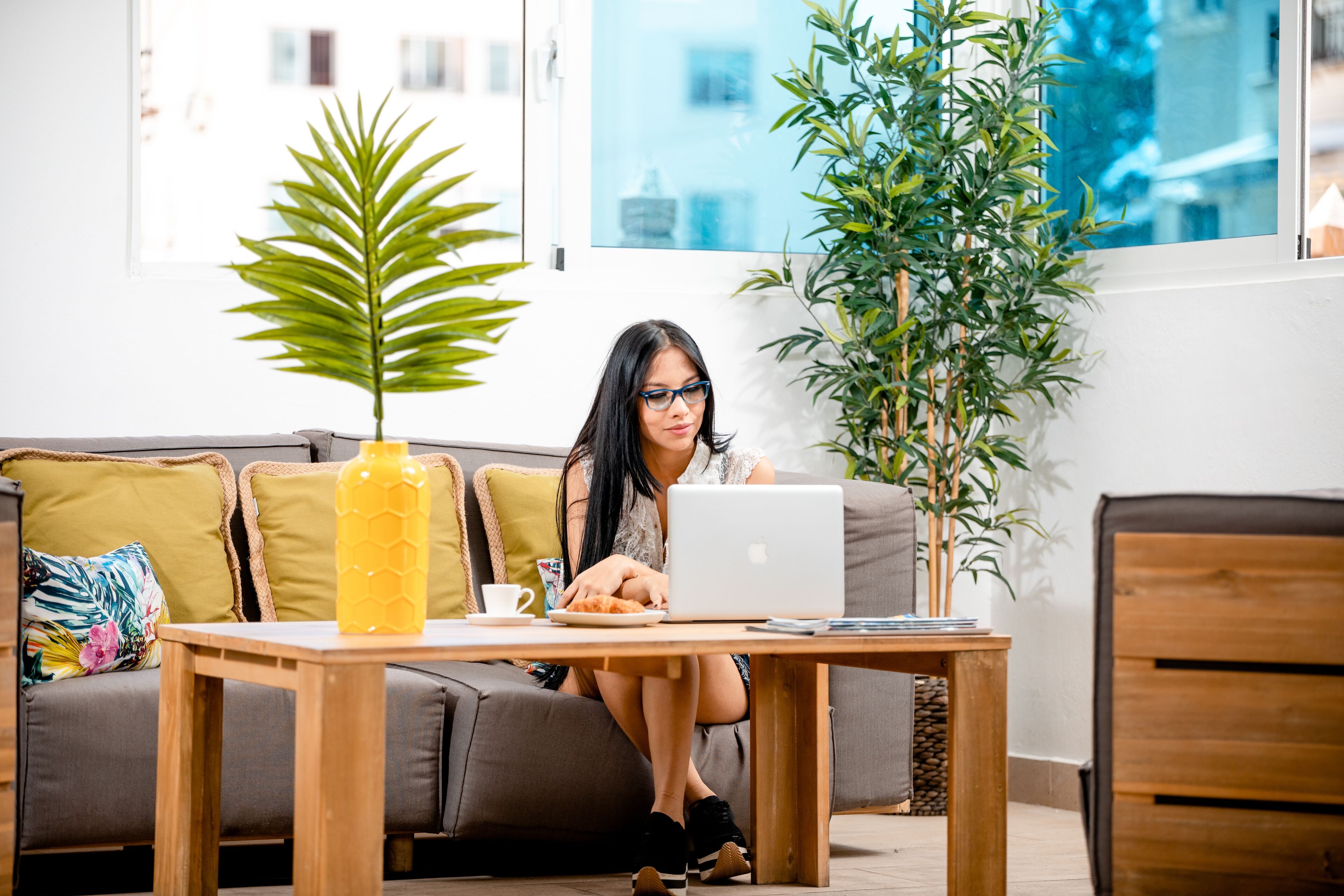 Uma pessoa sentada num sofá com um laptop, uma mesa de madeira com uma planta e uma garrafa amarela à frente.