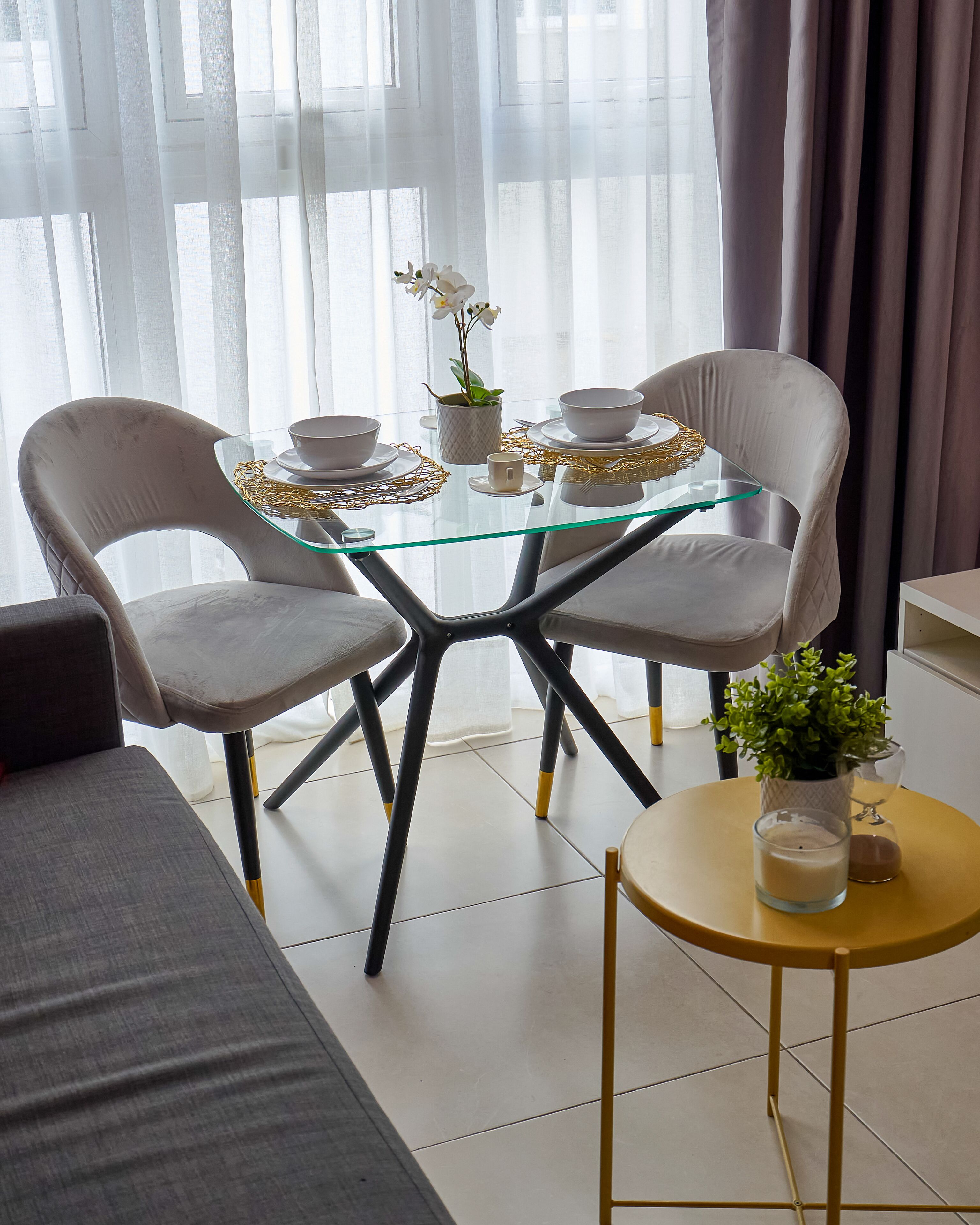 Uma área de refeições moderna com uma mesa de vidro, duas cadeiras, um vaso de planta e uma pequena mesa redonda com uma vela.