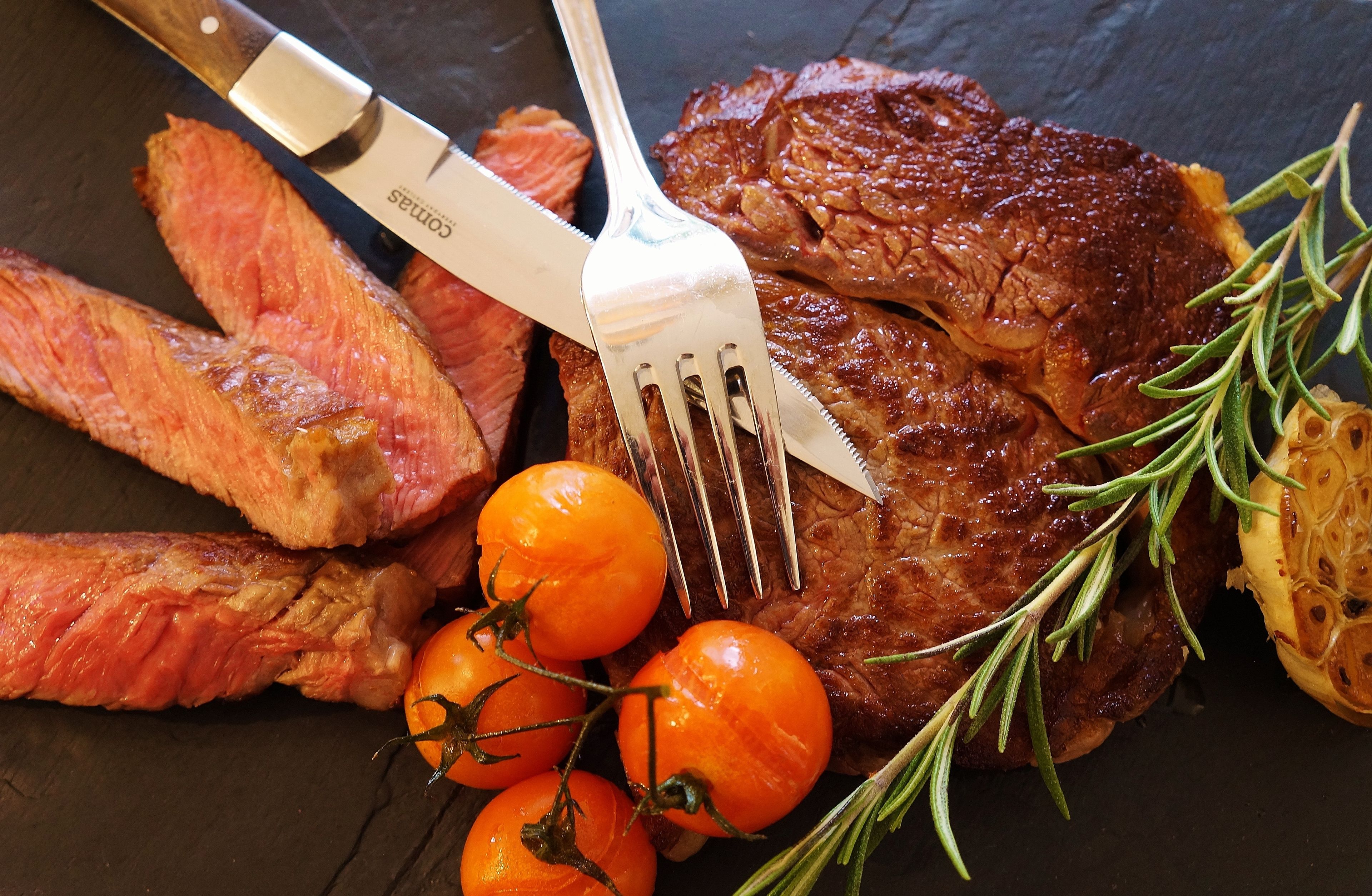Bife com garfo e faca em um prato, acompanhado de tomates cereja e alecrim.