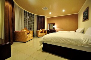 Quarto de hotel com uma cama grande, duas poltronas, uma escrivaninha e uma janela com cortinas.