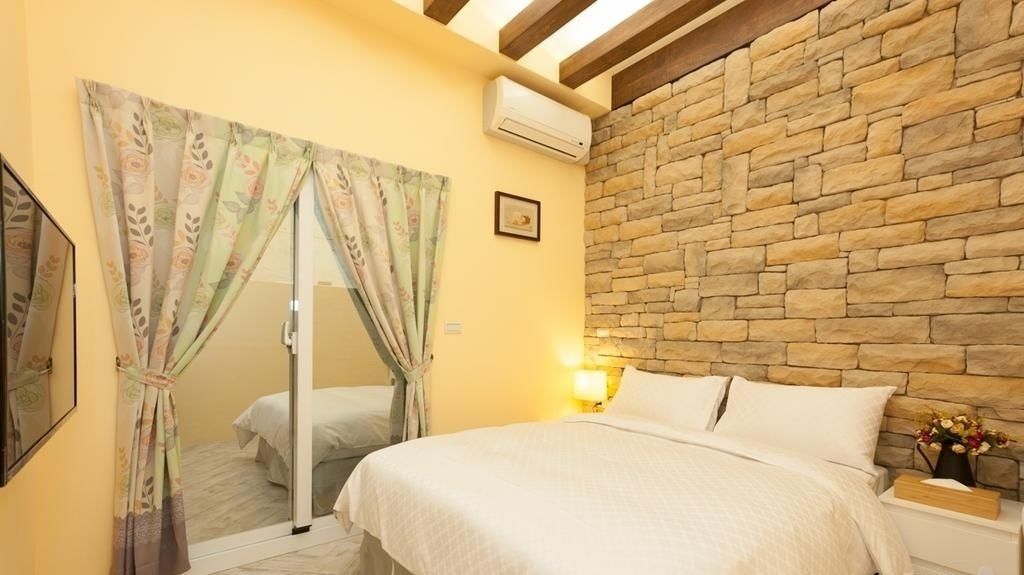 Quarto de hotel com parede de pedra, cama com roupa de cama branca, mesa de cabeceira com abajur e janela com cortinas.