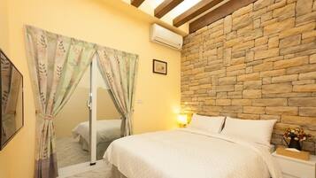 Quarto de hotel com parede de pedra, cama com roupa de cama branca, mesa de cabeceira com abajur e janela com cortinas.