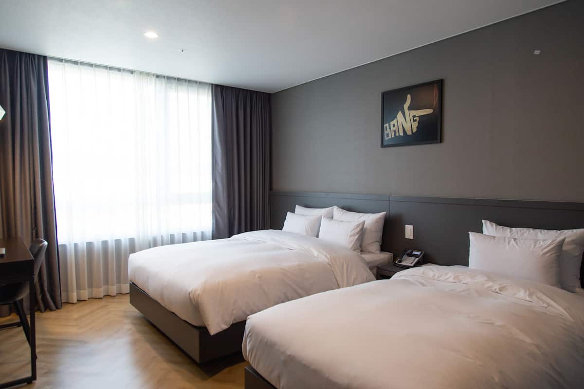 Quarto de hotel com duas camas, uma escrivaninha, uma cadeira, uma mesinha de cabeceira e um quadro na parede.