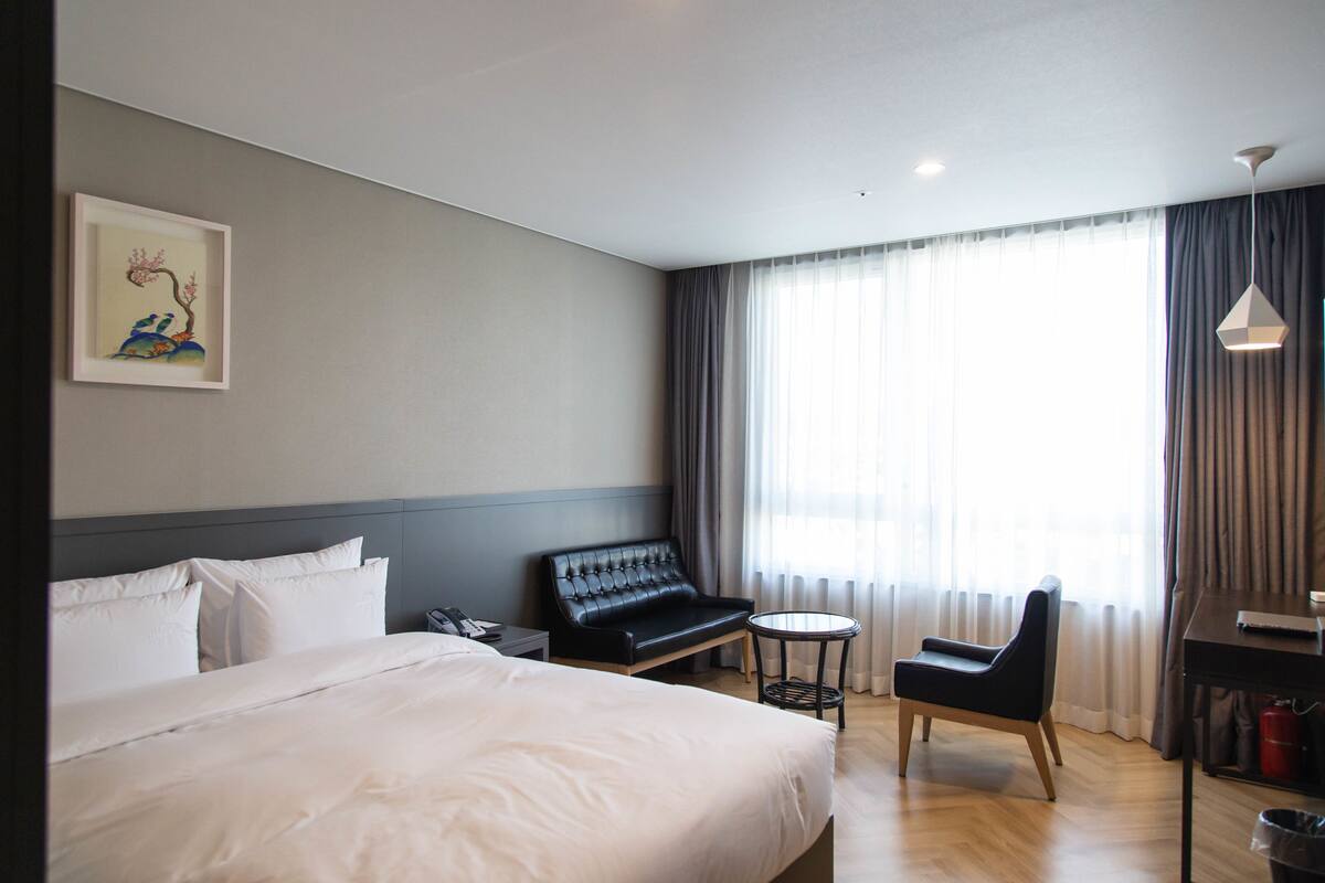 Quarto de hotel com uma cama grande, um sofá de couro preto, uma mesinha redonda, uma escrivaninha com cadeira e um quadro na parede.