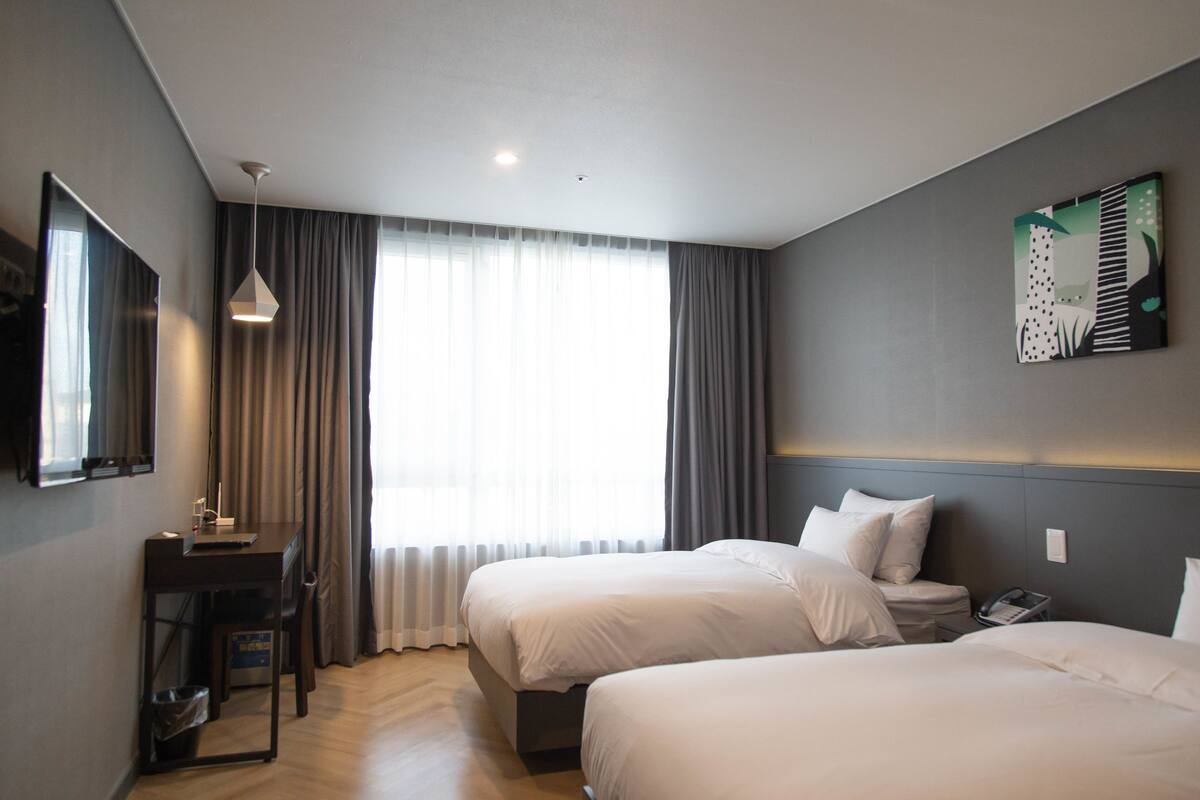 Quarto de hotel com duas camas, uma escrivaninha, uma cadeira, uma televisão e um quadro na parede.
