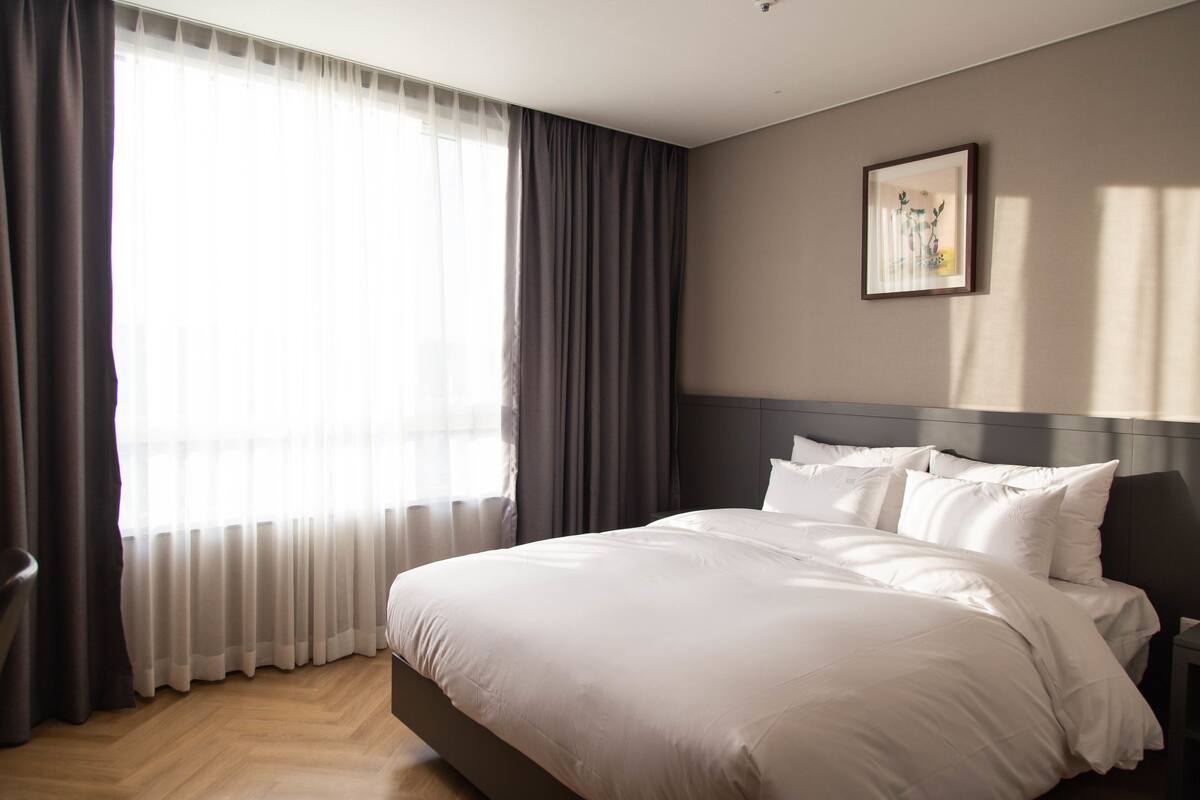 Quarto de hotel com uma cama grande, cabeceira, quadro emoldurado na parede e janela com cortinas.
