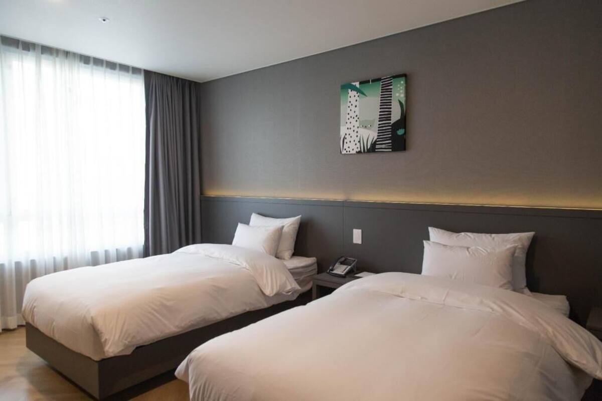 Quarto de hotel com duas camas, mesa de cabeceira, um quadro e janela com cortinas.