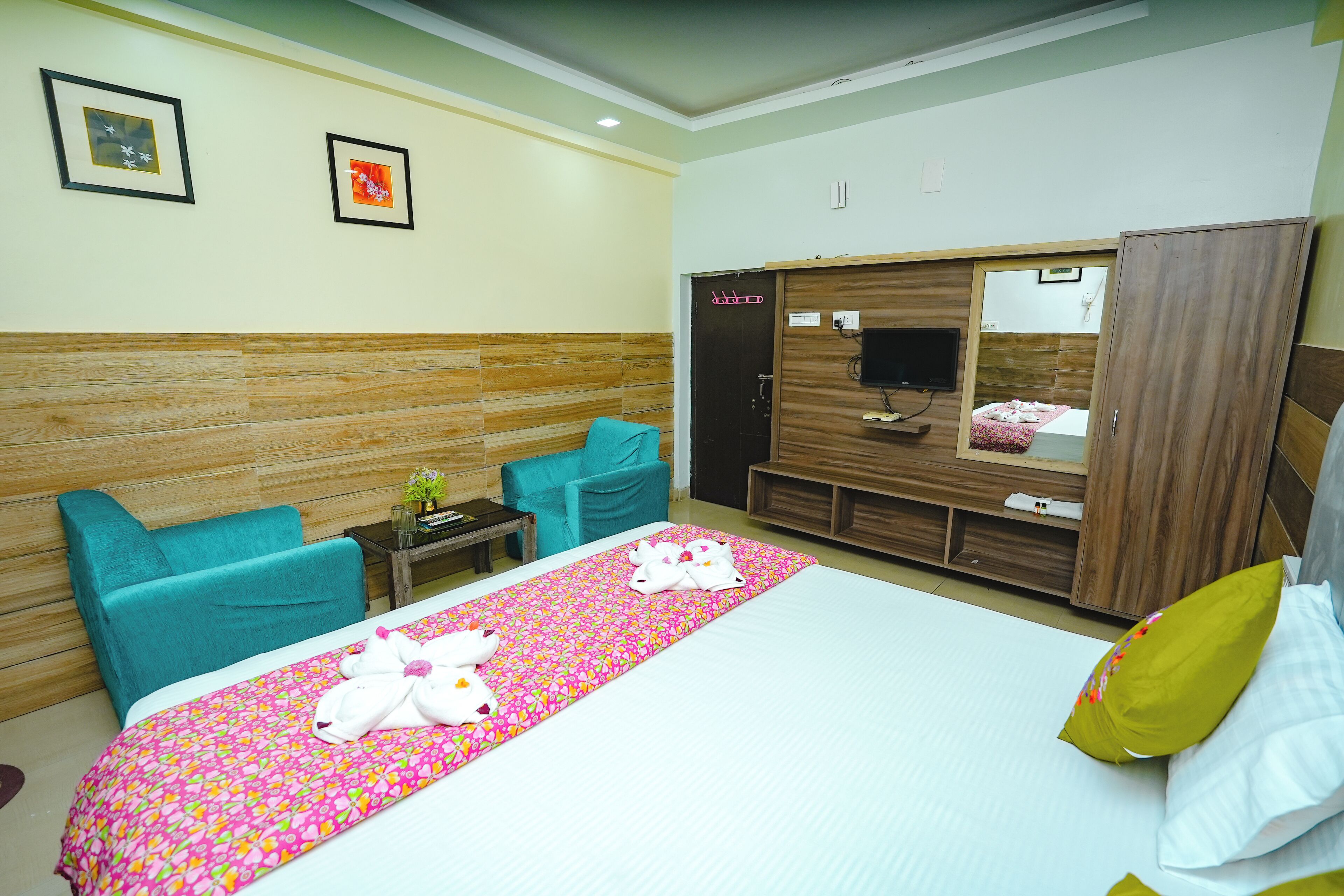 Quarto de hotel com uma cama, duas poltronas turquesa, um guarda-roupa de madeira, uma televisão e uma pequena mesa com uma planta.