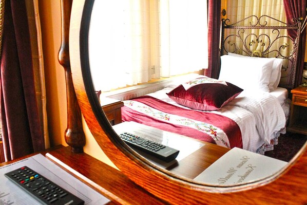 Quarto de hotel com escrivaninha de madeira, cama com colcha vermelha e branca, mesa de cabeceira e espelho.