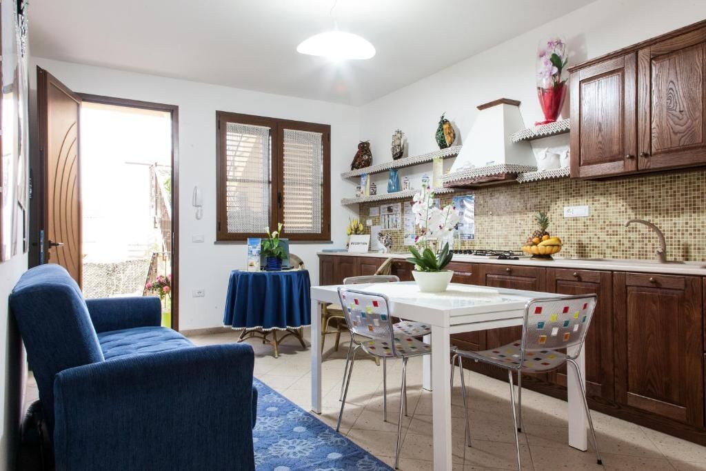 Cozinha com mesa de jantar, cadeiras e um sofá azul. Há armários, uma janela com persianas e painel de azulejos.