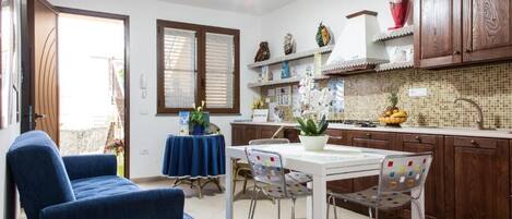 Cozinha com mesa de jantar, cadeiras e um sofá azul. Há armários, uma janela com persianas e painel de azulejos.