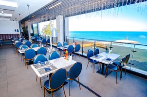 Restaurante com mesas e cadeiras dispostas para refeições, grandes janelas com vista para o mar e design de interiores moderno.