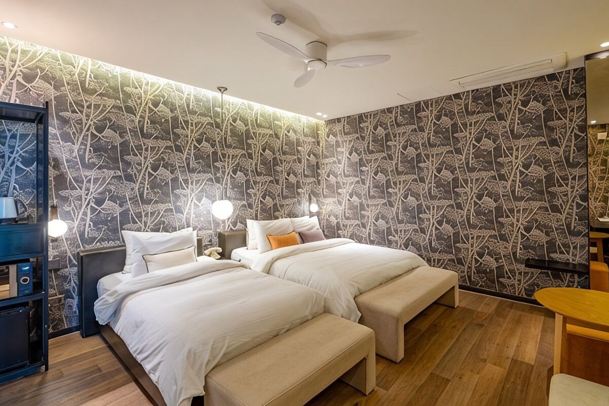 Quarto moderno com duas camas, uma mesinha de cabeceira, um banco, um ventilador de teto e uma pequena mesa de madeira.
