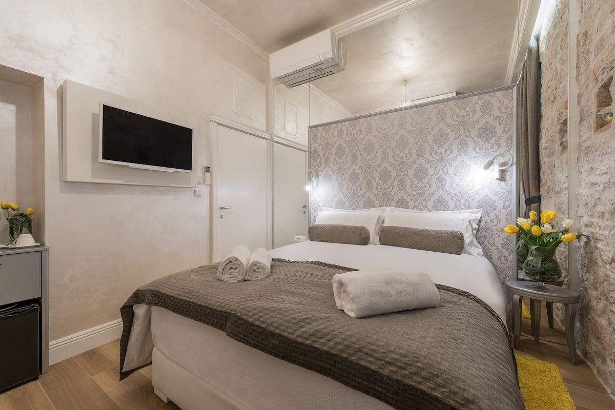 Um quarto moderno com uma cama grande, uma televisão de tela plana na parede, uma pequena mesa de cabeceira com um vaso de flores e um espelho decorativo.