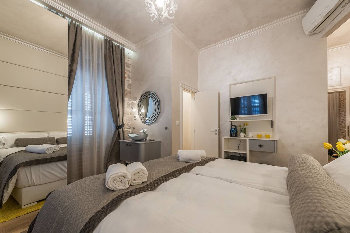 Quarto de hotel com duas camas, televisão, espelho, pequena mesa com abajur e prateleira com artigos de higiene pessoal.