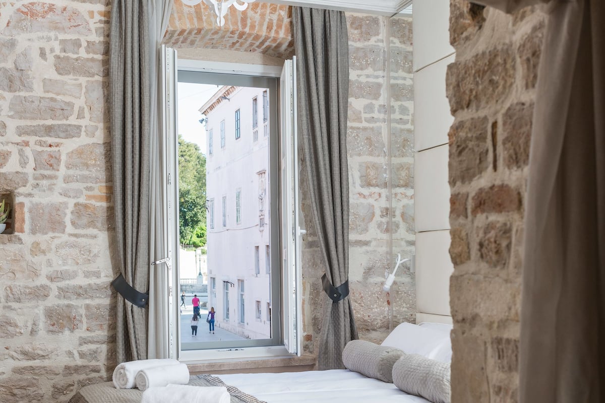 Quarto com cama, mesa de cabeceira com uma xícara e vista para a rua pela janela.