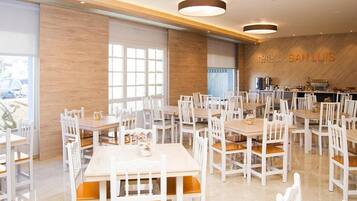 Restaurante de hotel com várias mesas e cadeiras, área de cozinha e um letreiro que diz "HOTEL SAN LUIS RESTAURANTE".