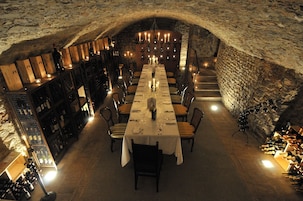 Uma área de refeições subterrânea com uma mesa comprida posta para uma refeição, cercada por estantes de vinho e paredes de pedra.