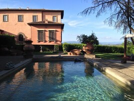Uma piscina límpida com borda de pedra, um edifício rosa com janelas e portadas, e um jardim bem cuidado com plantas em vasos e um pérgula.
