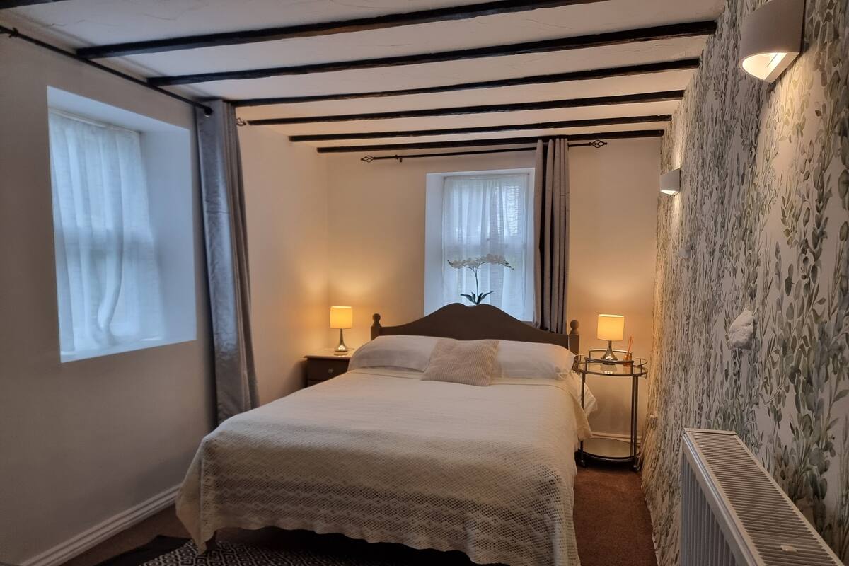 Quarto com uma cama grande, duas mesas de cabeceira com abajures, um radiador e uma janela com cortinas.