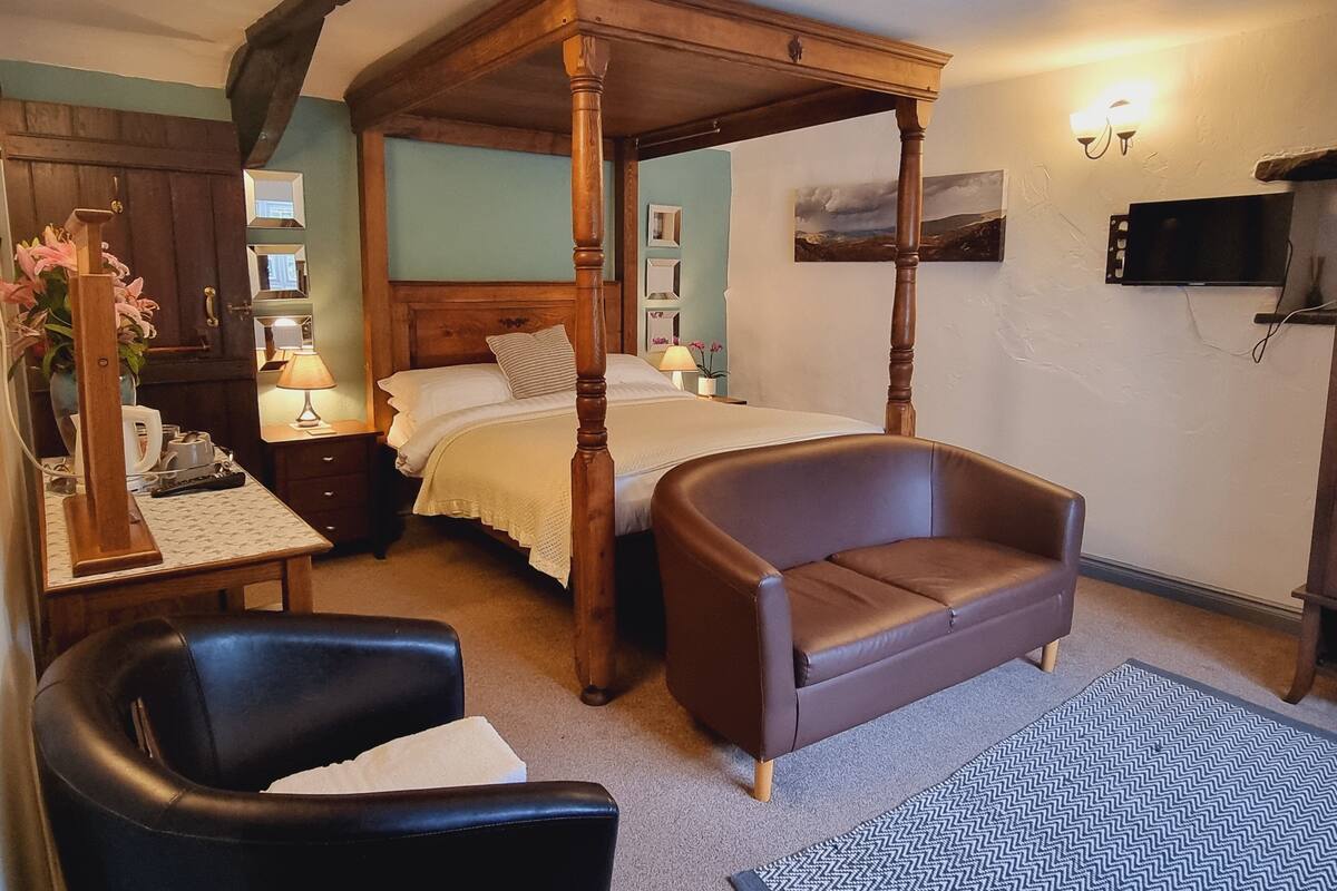 Uma cama de dossel com cabeceira de madeira, uma poltrona de couro marrom, uma mesa de cabeceira com abajur e uma televisão fixada na parede.