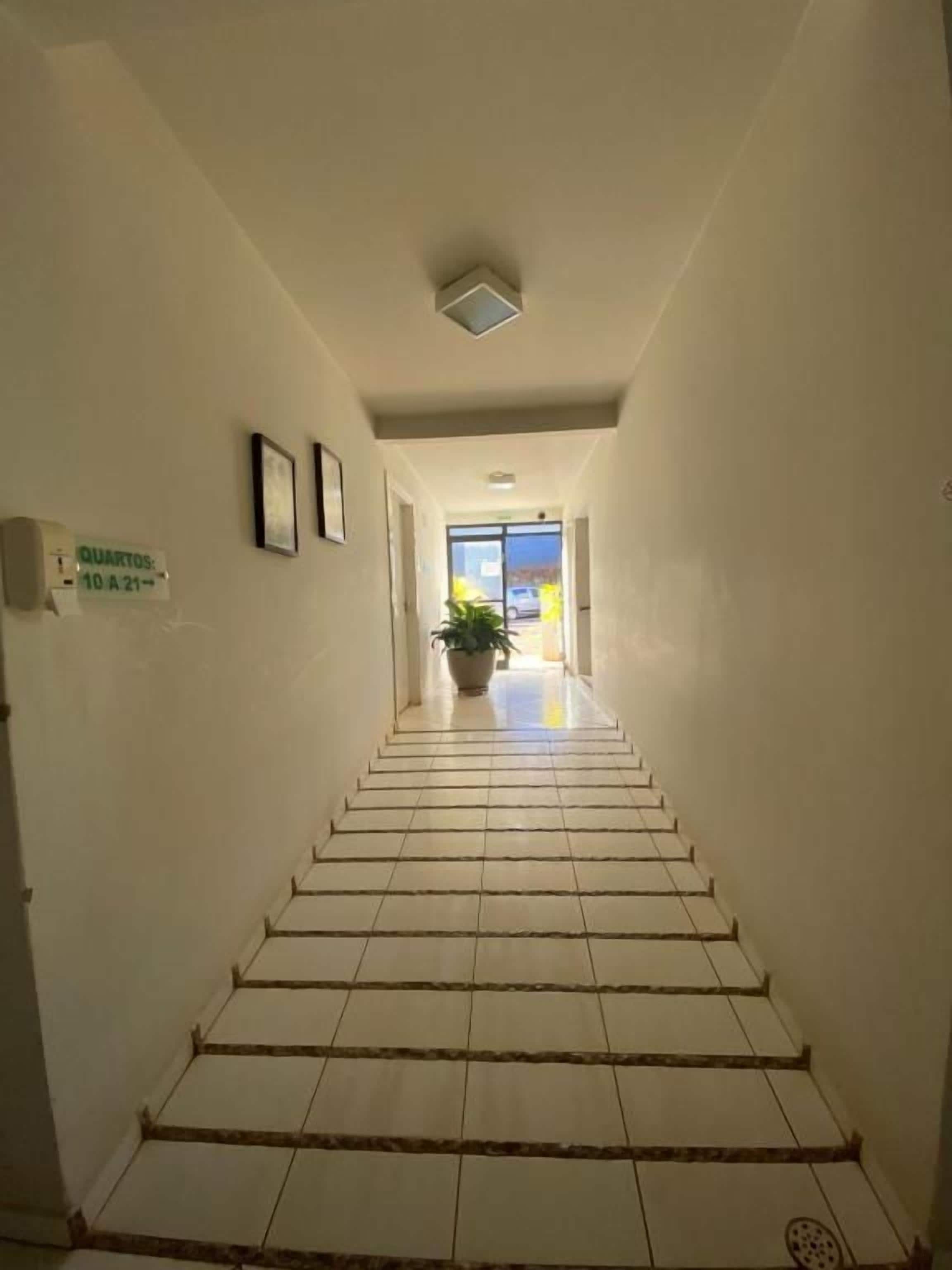 Corredor com piso em azulejo que leva a um espaço iluminado com uma planta.