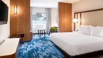 Quarto de hotel com uma cama grande, uma escrivaninha, uma cadeira, uma TV e uma janela com vista para árvores.