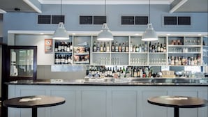 Uma área de bar com balcão, prateleiras de garrafas, copos e um cardápio. Há duas mesas redondas com bancos.