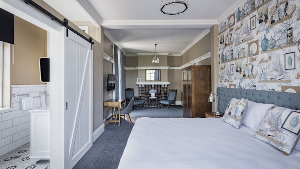 Quarto de hotel com uma cama grande, uma mesa com duas cadeiras, banheiro com pia e vaso sanitário brancos, e uma parede decorada com quadros.