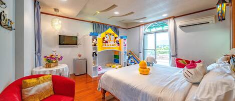 Quarto de hotel com uma cama, poltrona vermelha, casinha de brincar e televisão na parede.