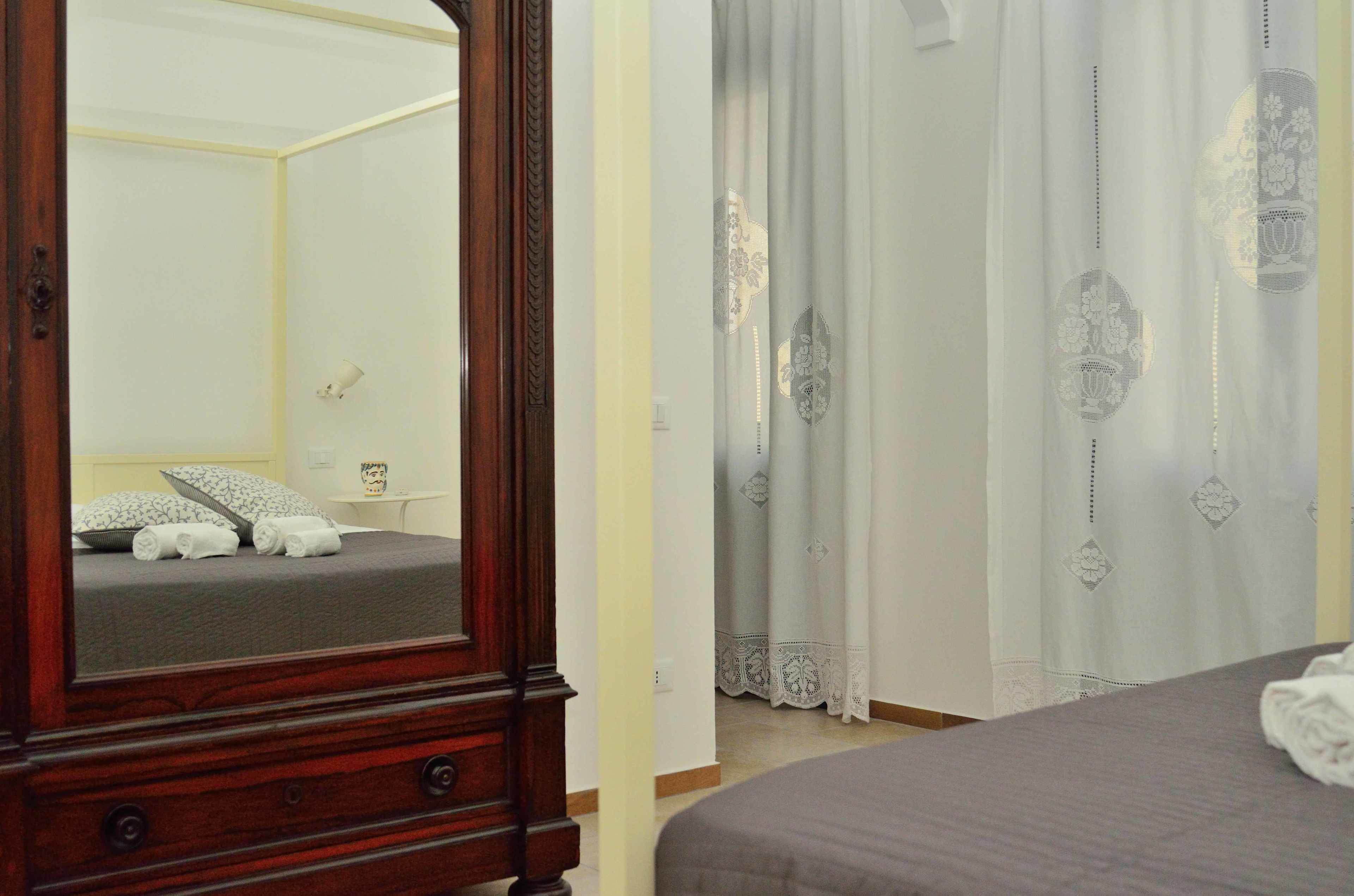 Quarto com um espelho grande, uma cama com colcha cinza e cortinas brancas com estampas florais.
