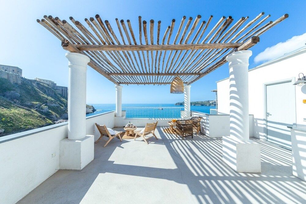 Um terraço no telhado com pérgola de madeira, colunas brancas e vista para o mar e uma fortaleza.
