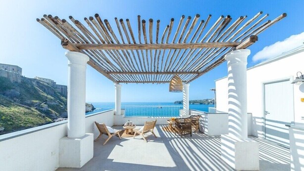 Um terraço no telhado com pérgola de madeira, colunas brancas e vista para o mar e uma fortaleza.