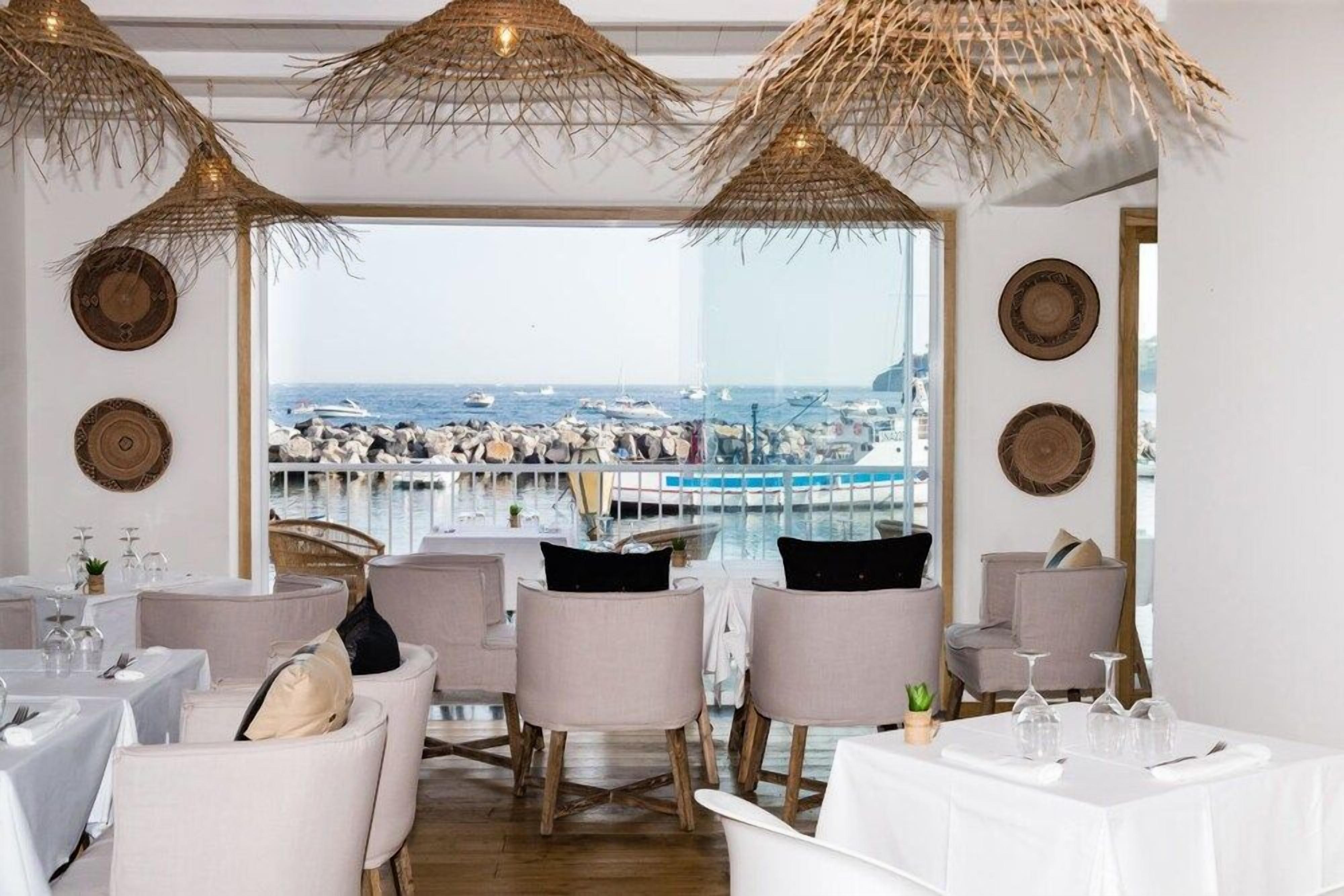 Um restaurante com mesas e cadeiras, vista para barcos e um porto de iates, e pratos decorativos na parede.