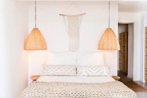 Quarto com cama, duas luminárias pendentes e um painel de macramê na parede.