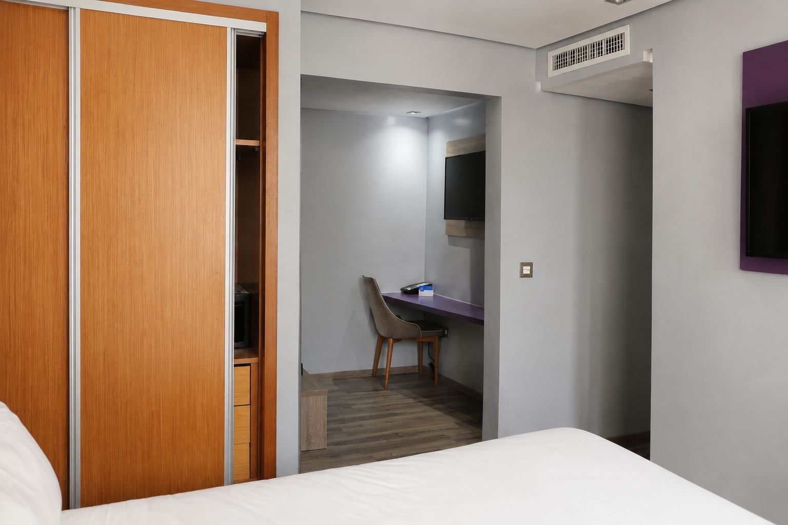 Quarto de hotel com cama, escrivaninha com cadeira, guarda-roupa e televisão na parede.