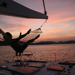 Uma pessoa relaxando numa rede no convés de um barco durante o pôr do sol.