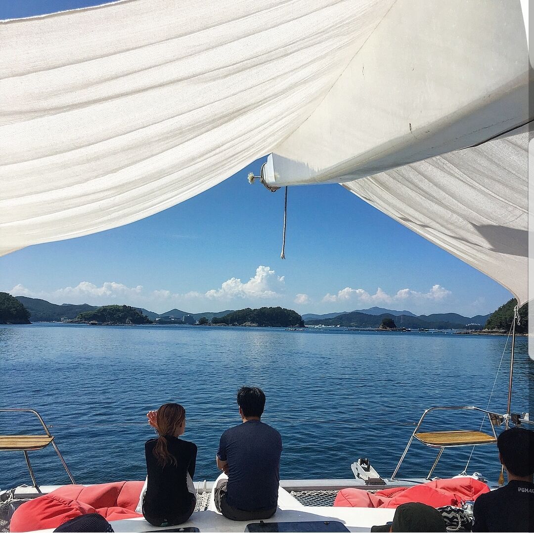 Um barco com toldo branco, duas pessoas sentadas e vista para águas calmas e terra ao longe.