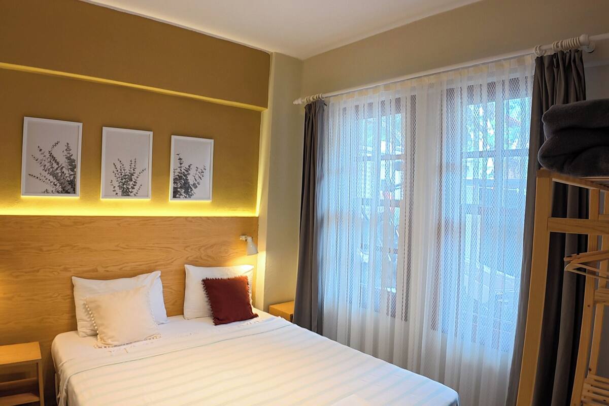 Um quarto de hotel espaçoso com uma cama grande, cabeceira de madeira, mesa de cabeceira e janela com cortinas translúcidas.