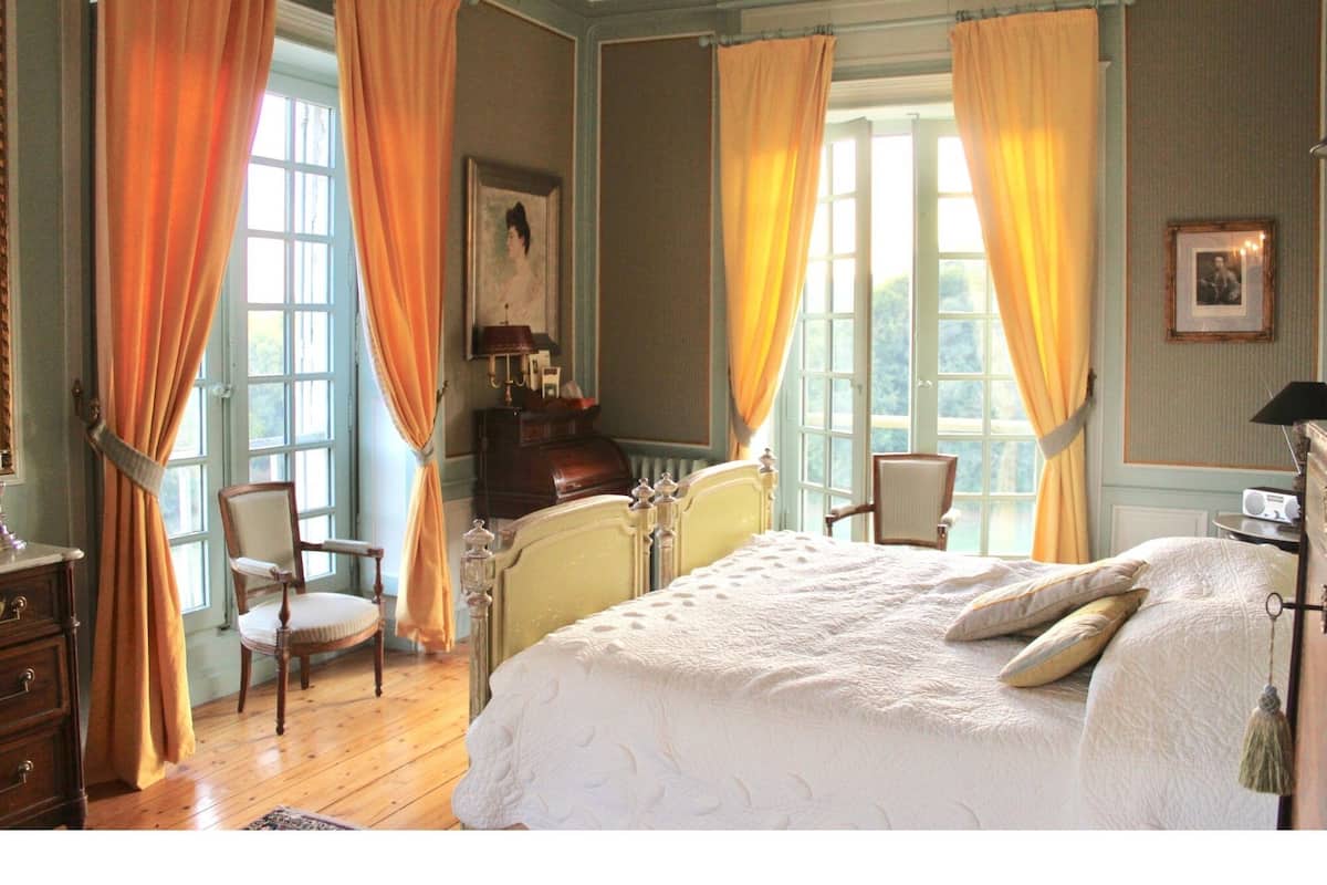 Quarto com cama grande, duas cadeiras, escrivaninha e janela com cortinas.