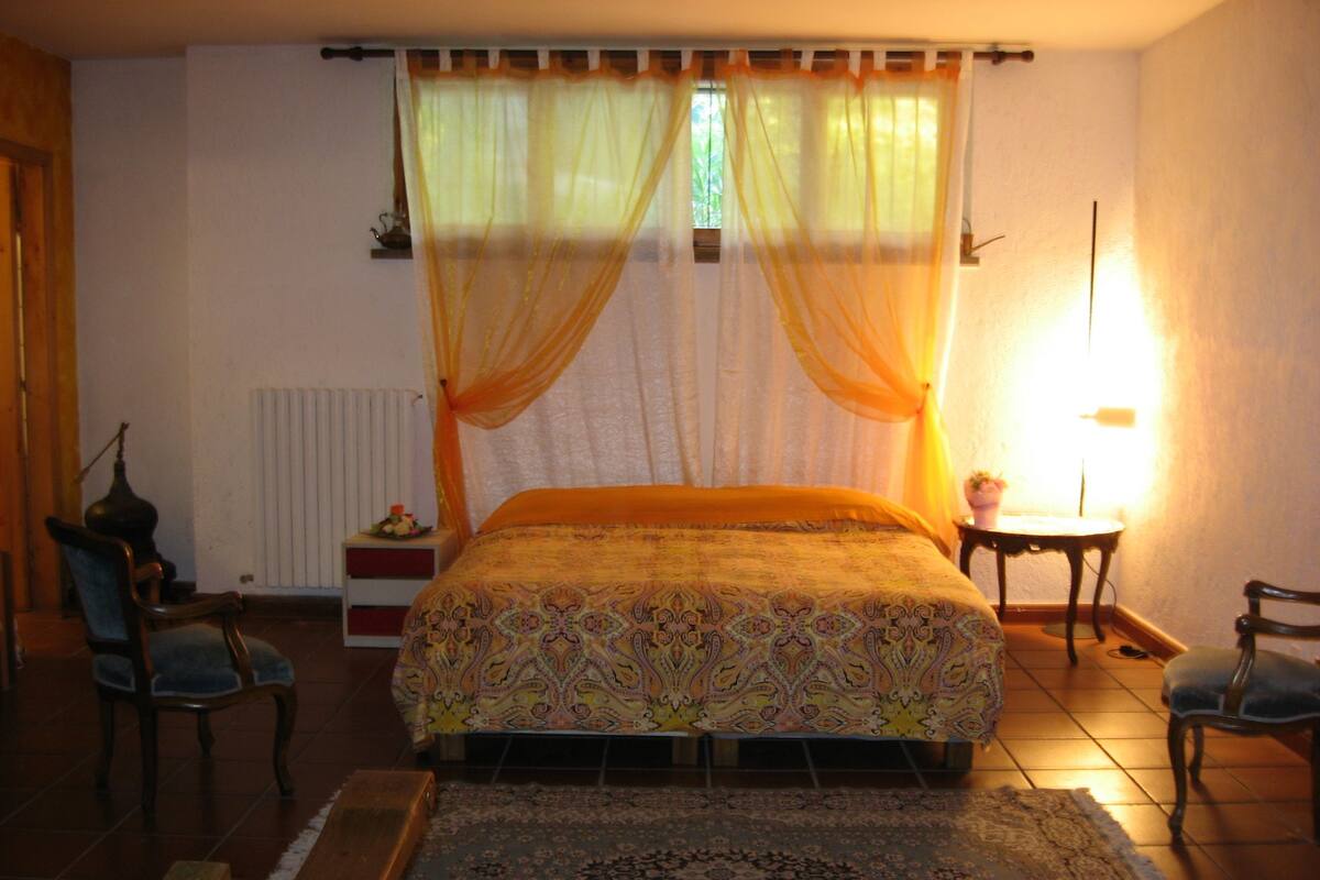 Quarto com cama, duas cadeiras, mesa de cabeceira, abajur e janela com cortinas translúcidas.