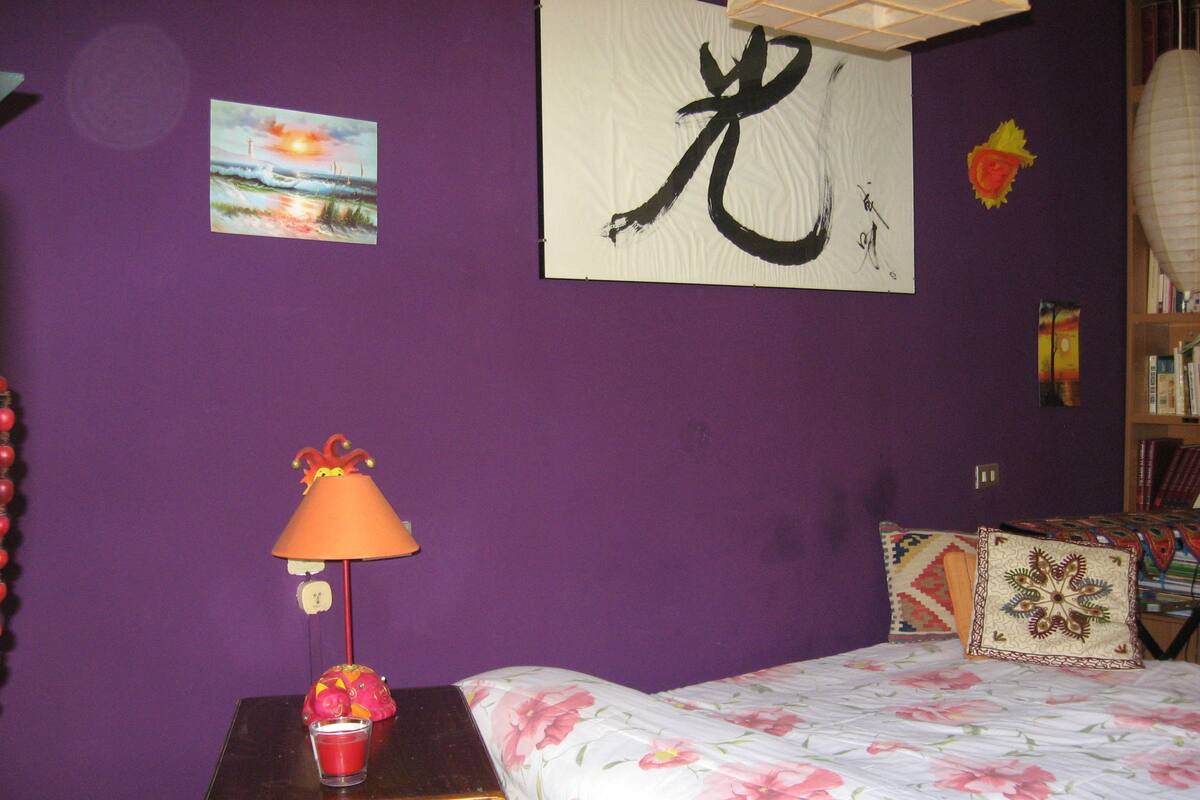 Quarto com parede roxa, cama com roupa de cama floral, mesa de cabeceira com abajur e estante de livros.
