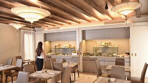 Uma sala de jantar moderna com vigas de madeira no teto, mesas postas com cadeiras e uma cozinha visível através de um painel de vidro.