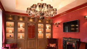 Um hall de entrada luxuoso com um lustre imponente, poltronas estofadas em vermelho, lareira e uma televisão grande fixada na parede.