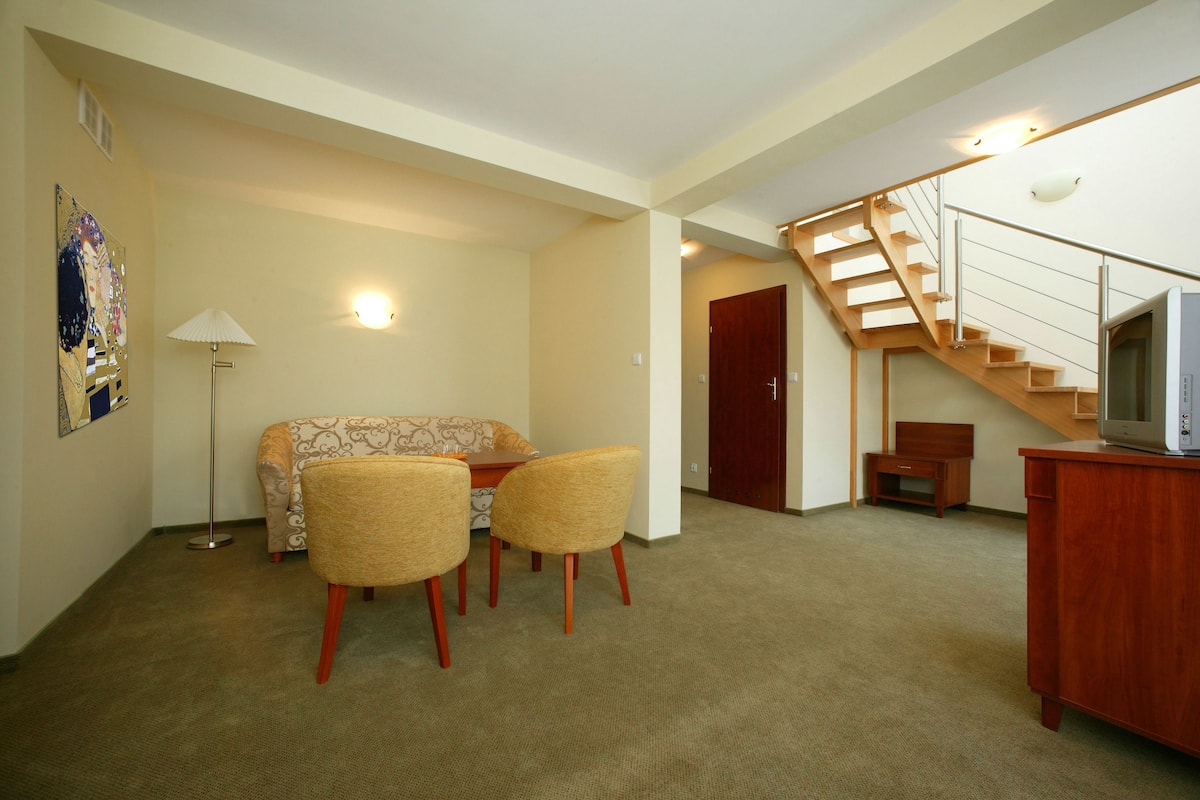 Uma sala de estar com sofá, duas cadeiras, mesa de centro, abajur, televisão e escada de madeira.