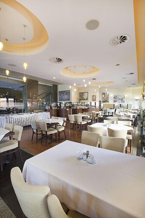 Um restaurante amplo com várias mesas e cadeiras, um espaço de bar e vista para a cidade através de grandes janelas.
