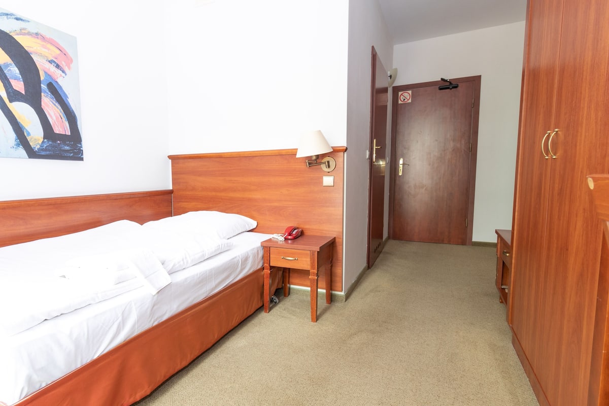 Uma cama de solteiro com cabeceira de madeira, uma mesa de cabeceira com abajur e um telefone. Uma porta com placa de "proibido fumar" é visível ao fundo.